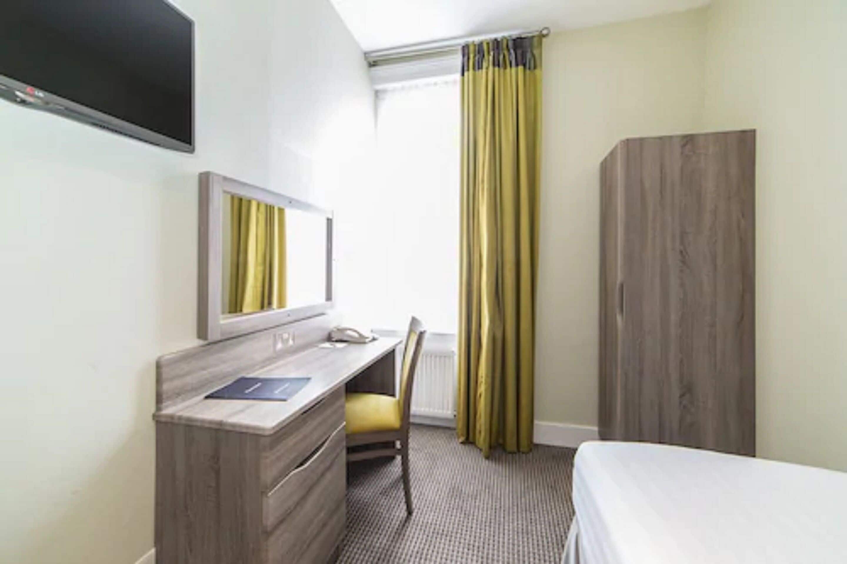 Quarto de hotel com cama, escrivaninha e cadeira, guarda-roupa, espelho e janela com cortinas.