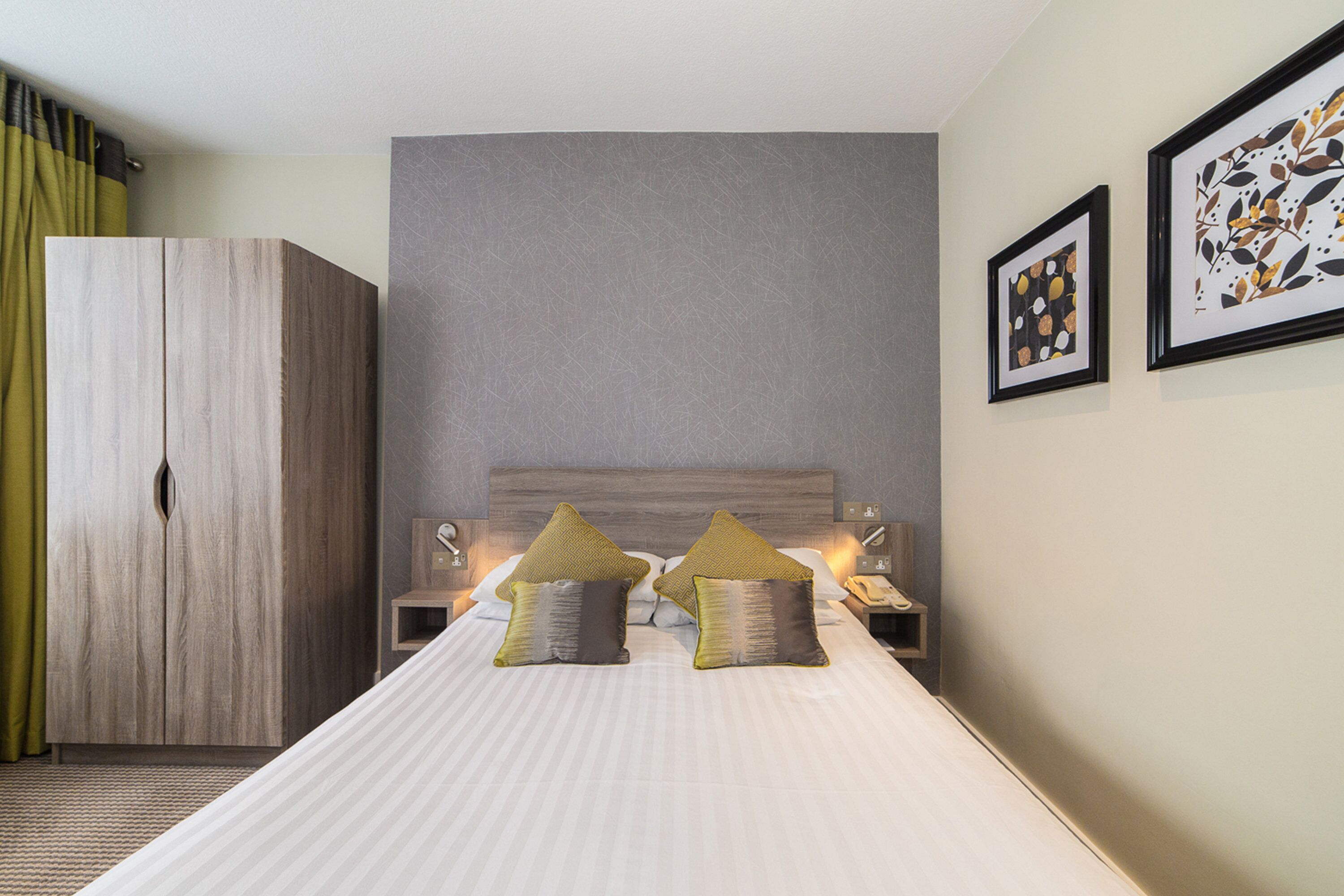Quarto de hotel com uma cama, duas mesas de cabeceira, um guarda-roupa e duas fotografias emolduradas na parede.