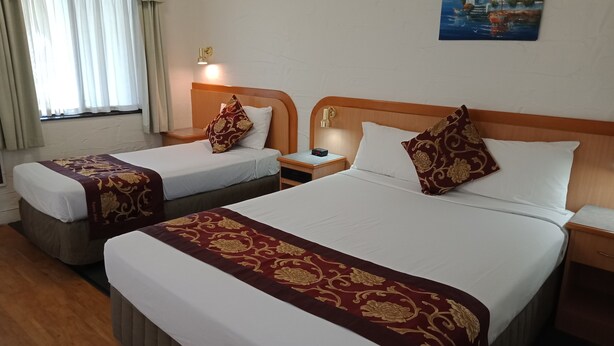 Quarto de hotel com duas camas, cada uma com cabeceira de madeira, roupa de cama branca e colcha com padrão dourado e vermelho. Há uma mesinha de cabeceira com abajur, uma janela com cortinas e um quadro com paisagem costeira na parede.