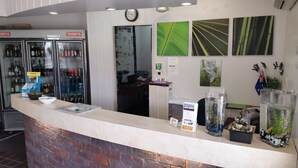 Uma área de recepção com balcão de tijolos, uma geladeira abastecida com bebidas e um relógio na parede.
