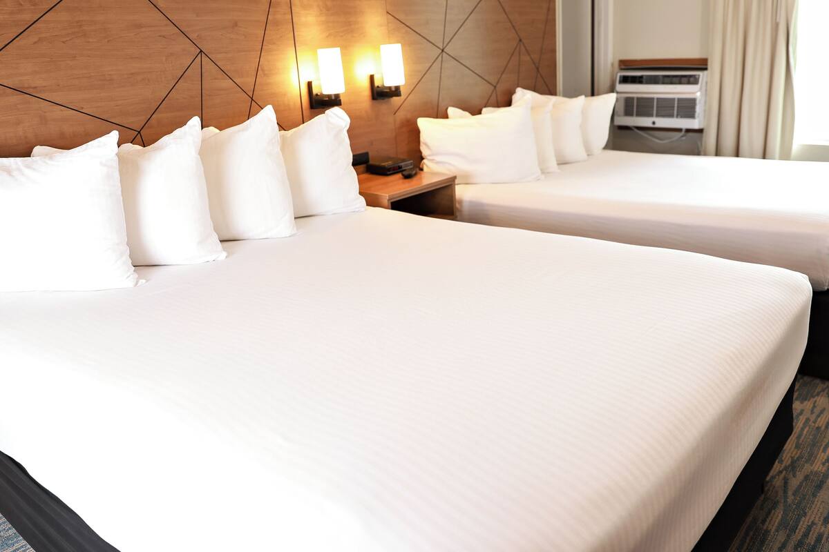 Quarto de hotel com duas camas, cabeceira de madeira, luminárias fixadas na parede, mesa de cabeceira e janela com cortinas.