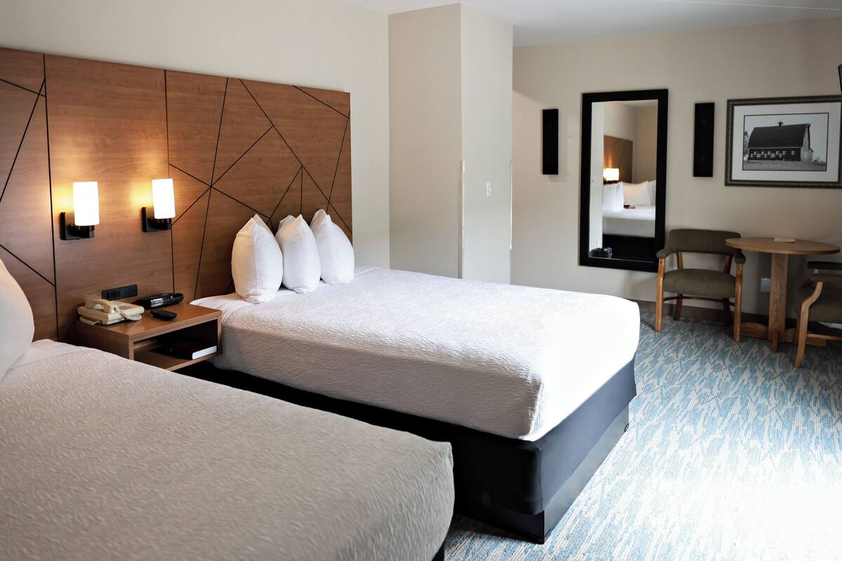 Quarto de hotel com duas camas, cabeceira de madeira, abajures, escrivaninha com cadeira, espelho e um quadro na parede.