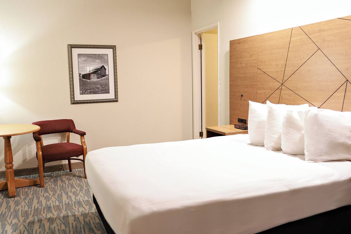 Quarto de hotel com cama, uma cadeira, uma mesinha, criado-mudo, quadro emoldurado e cabeceira de madeira.