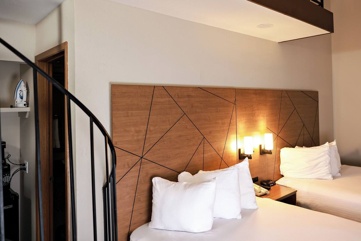 Quarto de hotel com cabeceira de madeira, duas camas com roupa de cama branca, mesa de cabeceira com abajur e escada com corrimão.