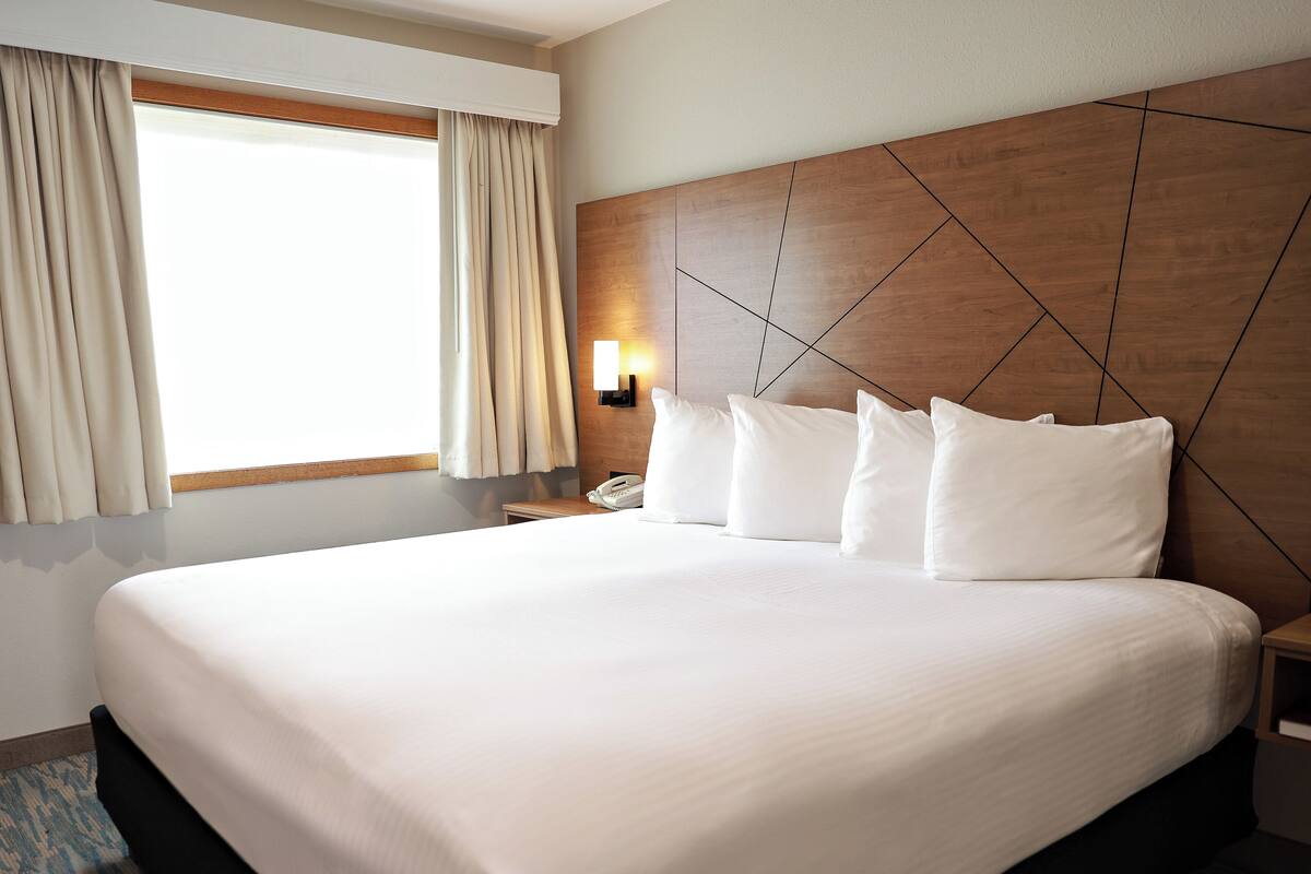 Quarto de hotel com uma cama grande, cabeceira de madeira, mesa de cabeceira com abajur e uma janela com cortinas.