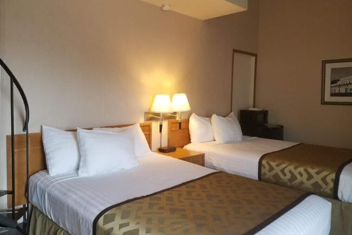 Quarto de hotel com duas camas, mesa de cabeceira com luminárias, quadro decorativo na parede e escada.