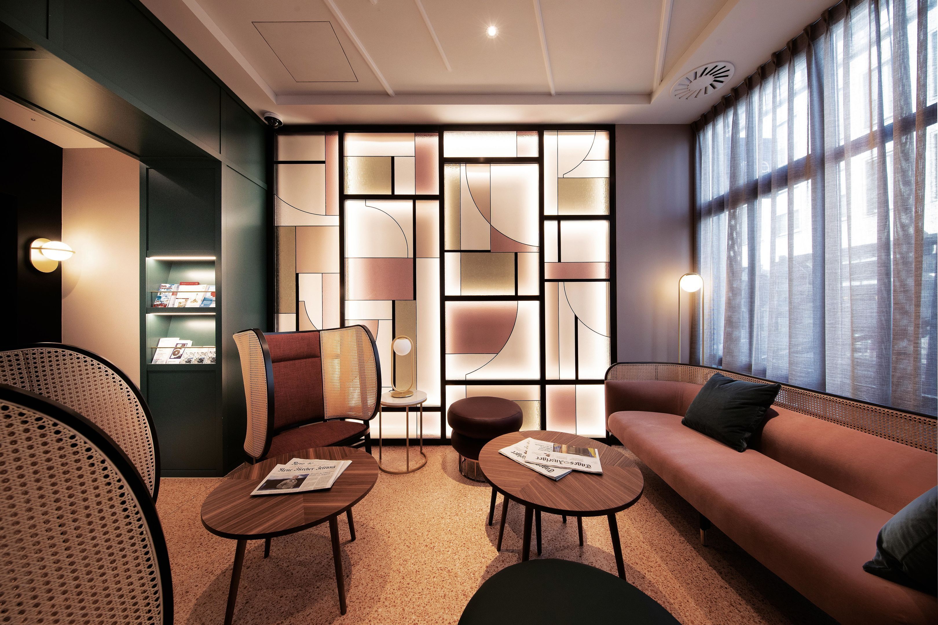 Quarto de hotel moderno com sofá, duas mesas redondas, uma cadeira e um painel decorativo na parede.