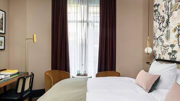 Quarto de hotel com cama, duas cadeiras, escrivaninha com abajur e janela com cortinas.