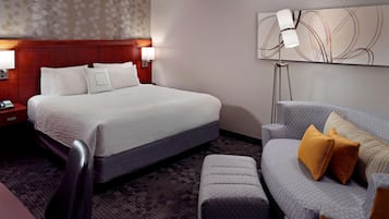 Quarto de hotel com uma cama grande, um sofá cinzento, uma mesinha e uma luminária na parede.