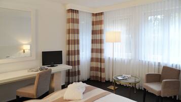 Quarto de hotel com cama, escrivaninha com computador, cadeira, mesa redonda com abajur e cortinas.