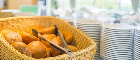 Uma cesta de vime cheia de pães e um par de pinças, ao lado de pratos brancos empilhados.