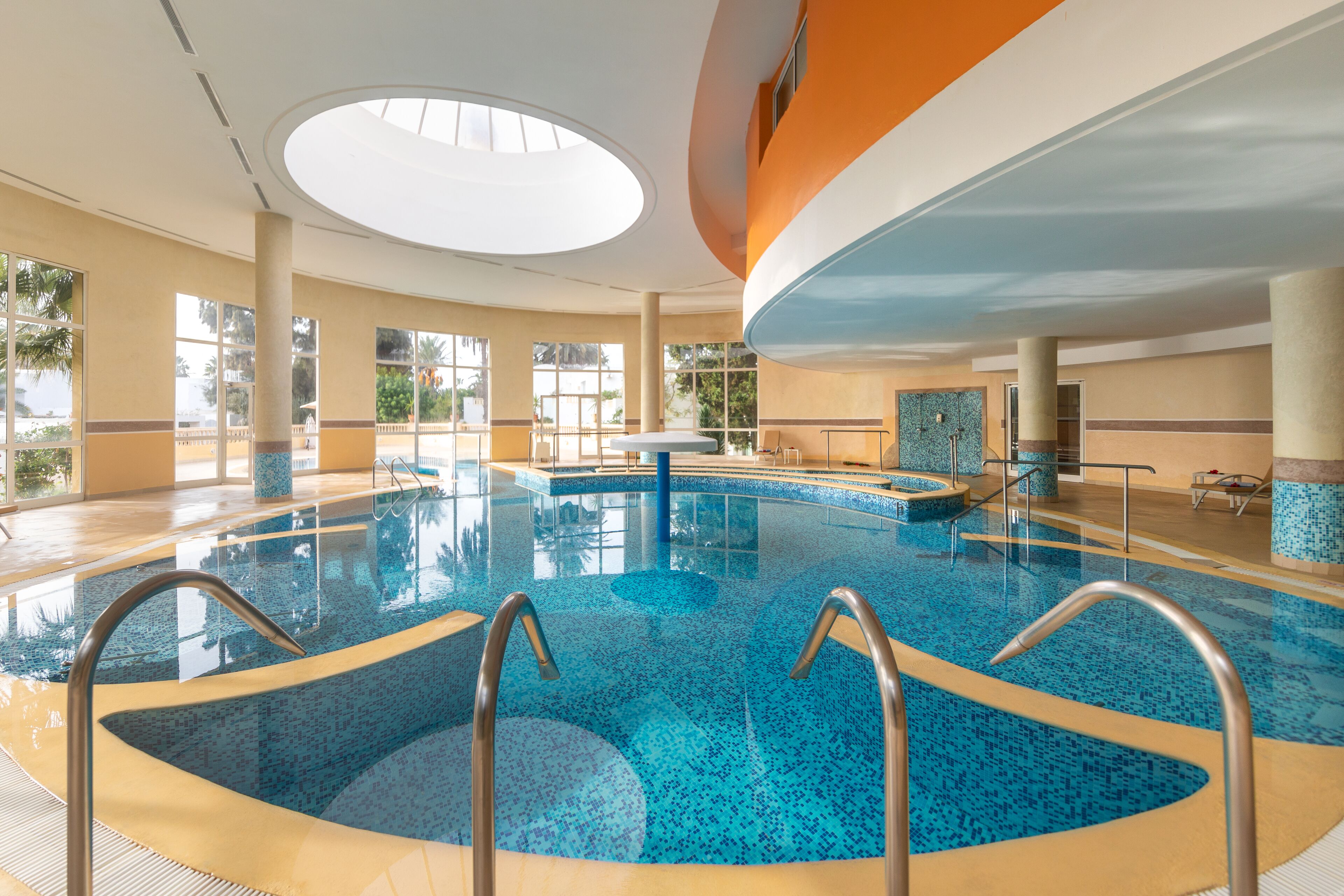 Uma piscina interior ampla com água azul cristalina, rodeada por paredes bege e grandes janelas.