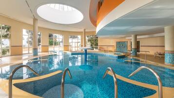 Uma piscina interior ampla com água azul cristalina, rodeada por paredes bege e grandes janelas.