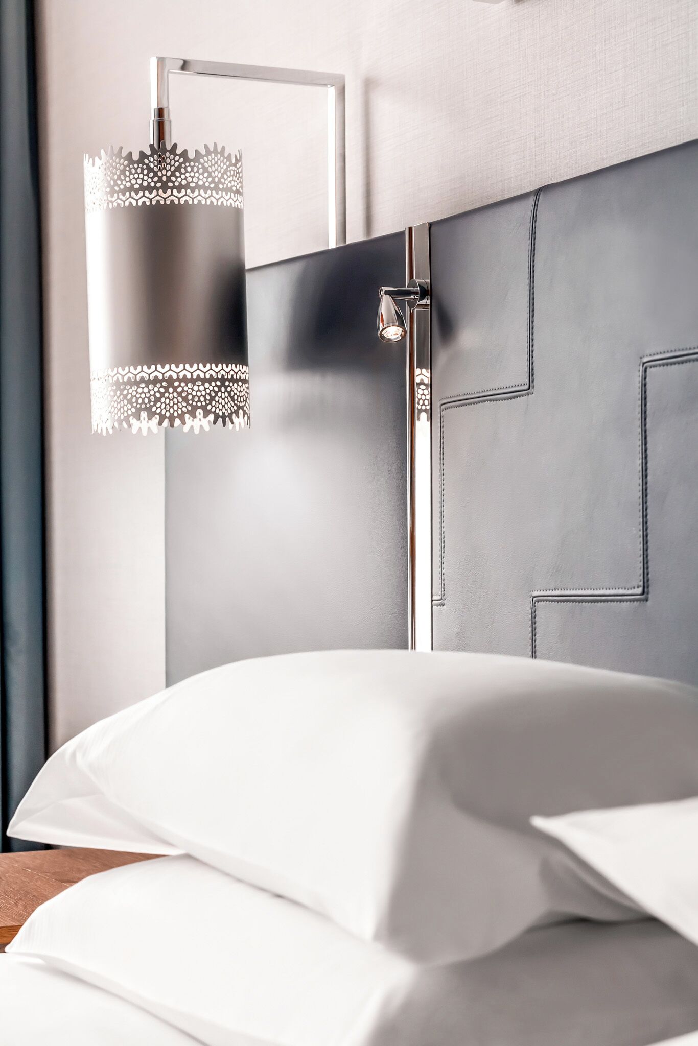 Quarto moderno com cabeceira cinza, luminária de mesa com cúpula estampada e travesseiros brancos bem organizados.