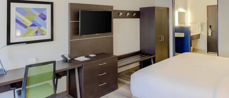 Quarto de hotel com cama, secretária com cadeira, televisão, guarda-roupa e casa de banho visível através de uma porta aberta.