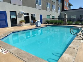 Uma piscina retangular com água azul cristalina, rodeada por um deck de concreto. Há uma escada de metal que leva à piscina, e uma placa indicando uma profundidade de 1,68 metros. A piscina fica ao lado de um edifício com várias janelas e uma porta, e uma pequena área coberta com uma cadeira e plantas em vasos.
