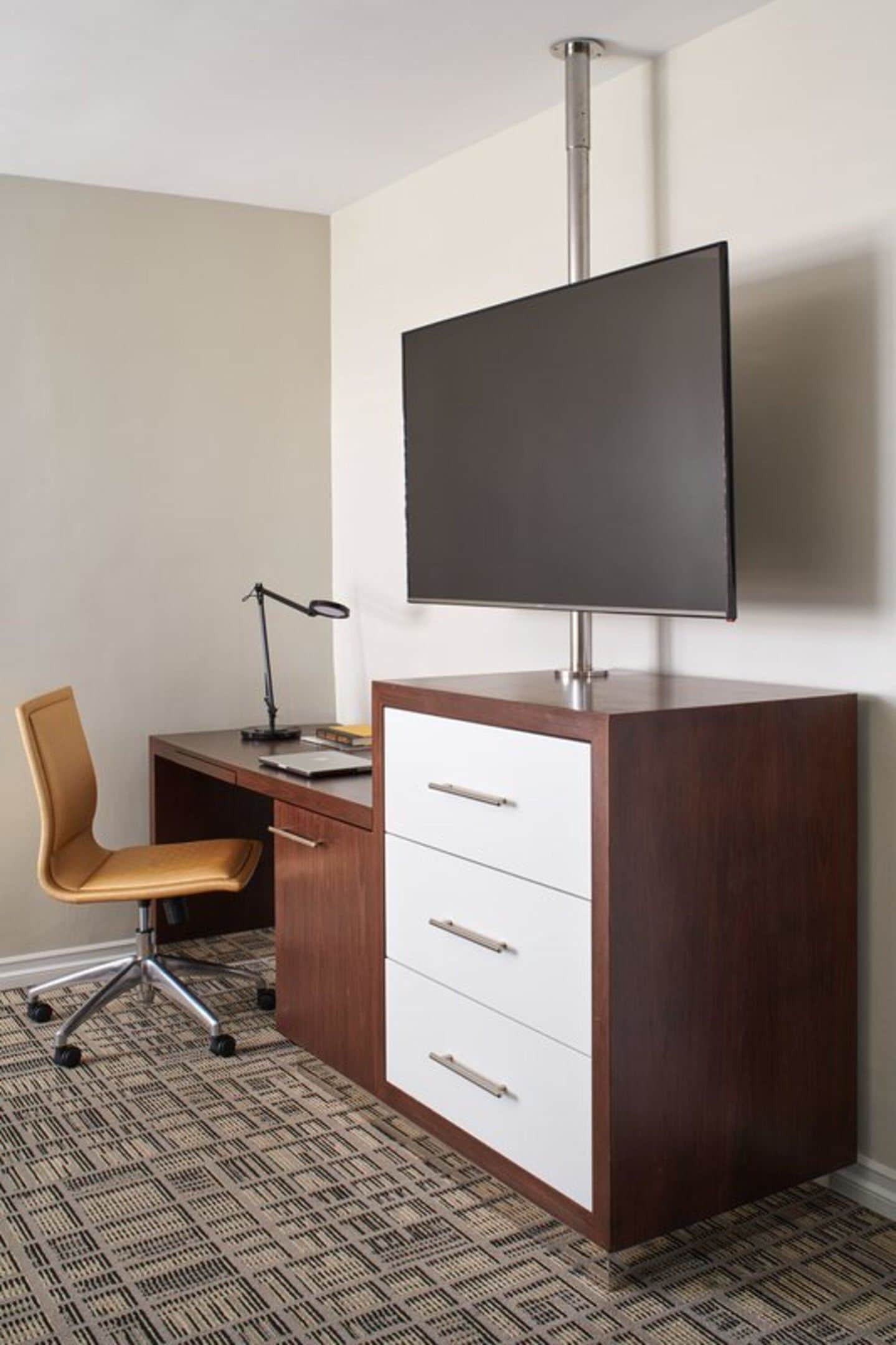 Quarto de hotel moderno com escrivaninha de madeira, uma cadeira, TV de tela plana na parede e carpete estampado.