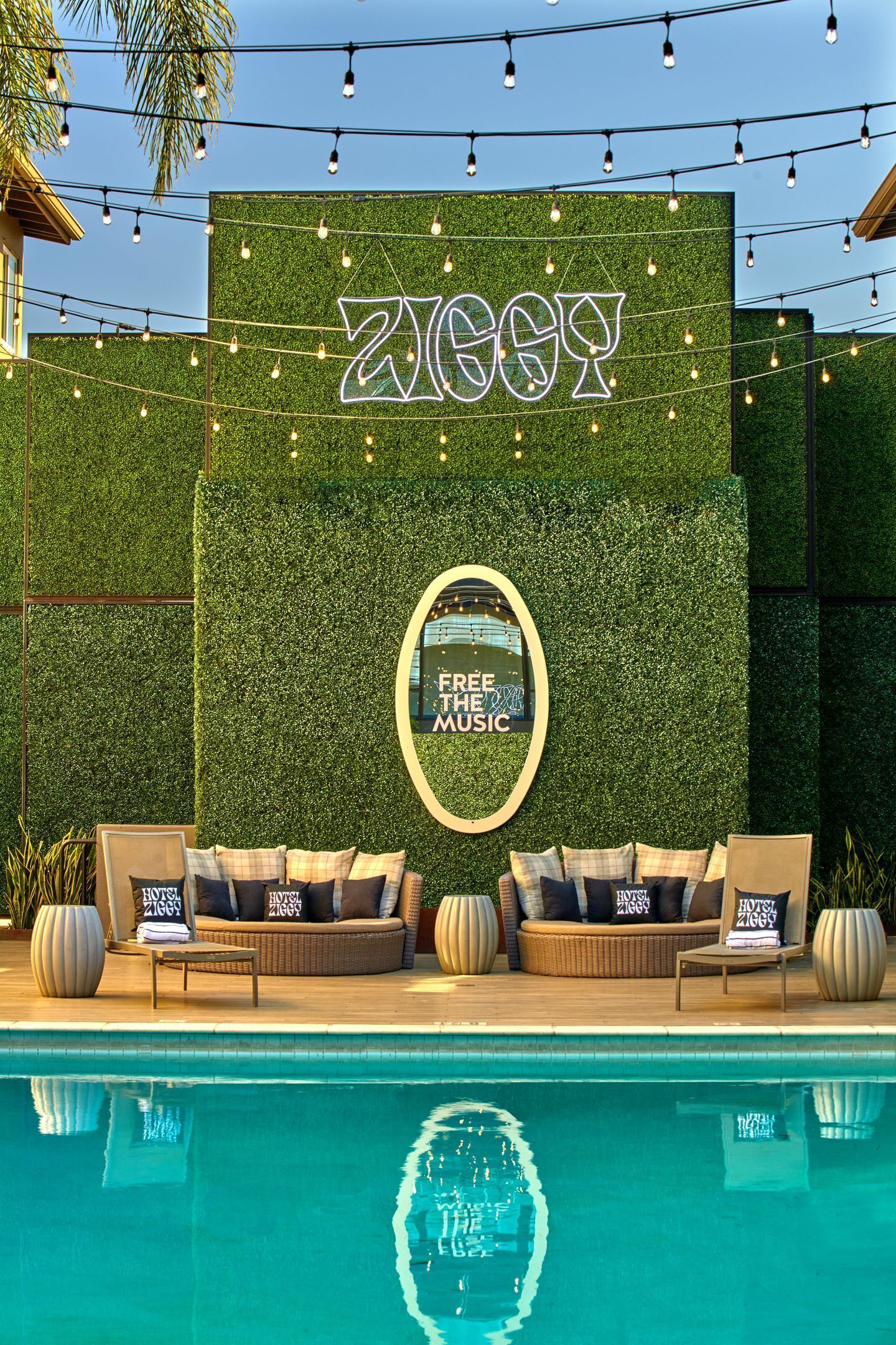 Uma área à beira da piscina com uma parede verde, uma placa com a inscrição 'FREE HEAR MUSIC' e assentos de lounge com almofadas rotuladas 'HOTEL ZIGZAG'.