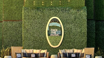 Uma área à beira da piscina com uma parede verde, uma placa com a inscrição 'FREE HEAR MUSIC' e assentos de lounge com almofadas rotuladas 'HOTEL ZIGZAG'.