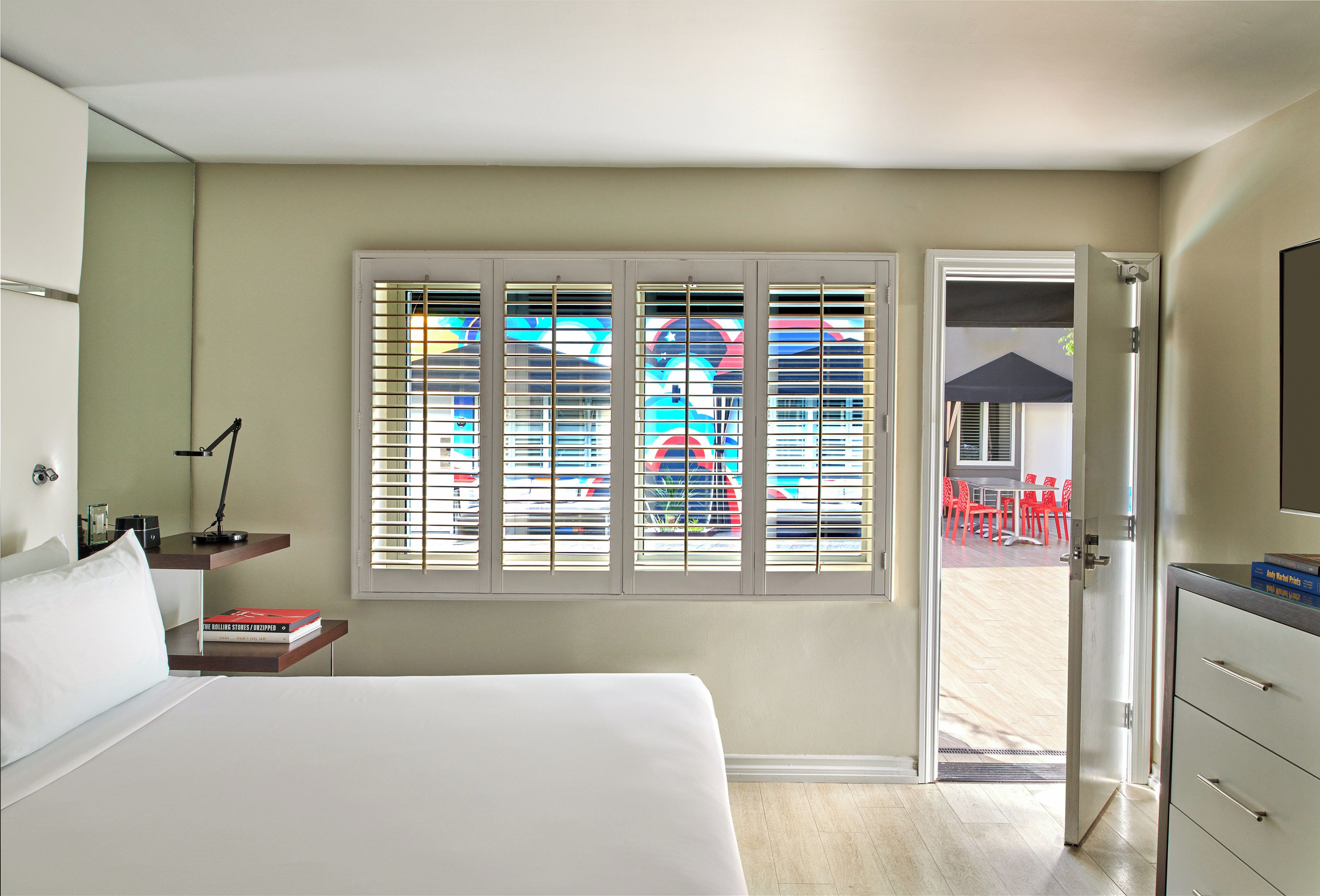 Quarto de hotel com cama, mesa de cabeceira, luminária e janela com persianas.