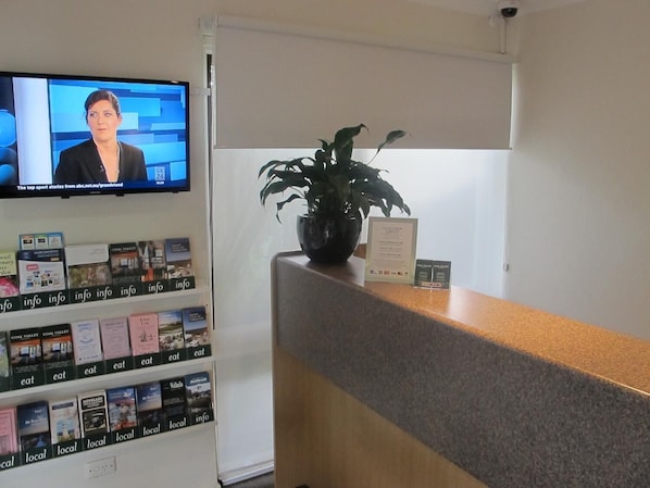 Uma área de recepção com televisão exibindo um canal de notícias, uma planta e folhetos informativos na parede.