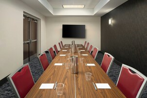Uma sala de conferências com uma longa mesa de madeira, cadeiras vermelhas, uma grande TV de tela plana e um projetor na parede.