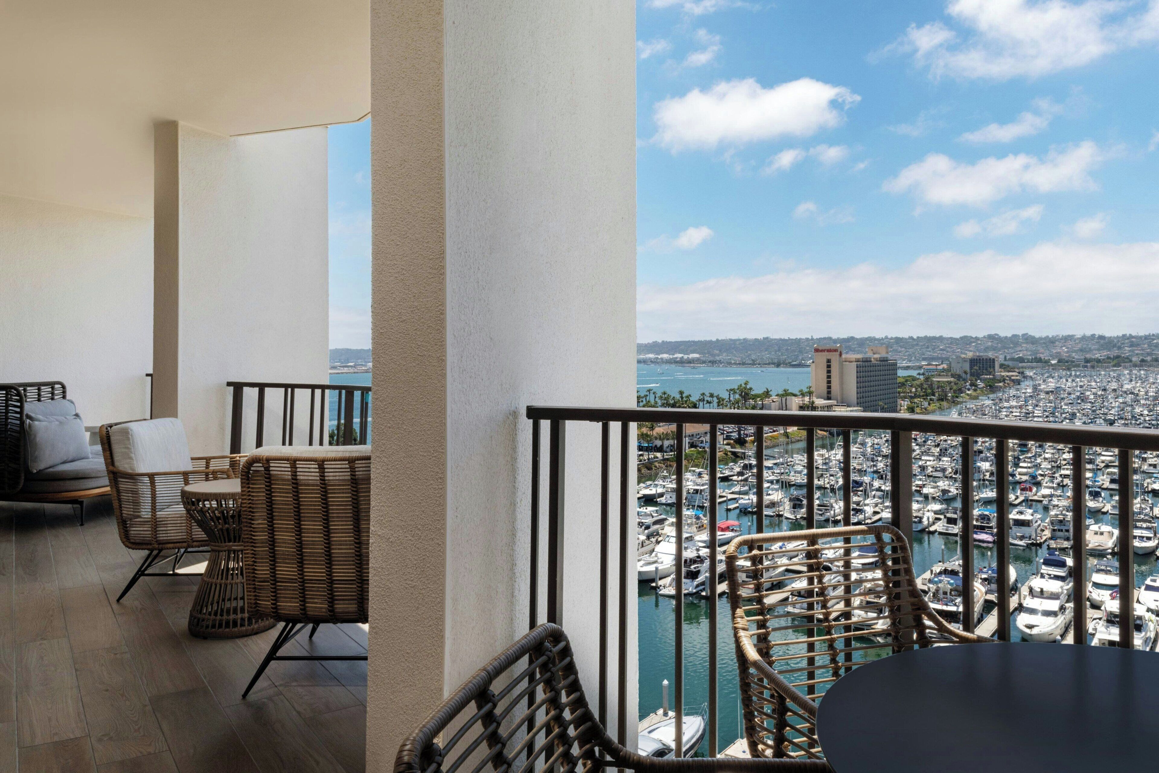 Uma varanda com móveis de vime, vista para uma marina e um horizonte urbano ao fundo.