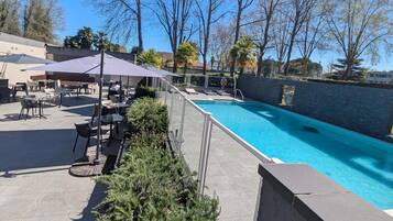 Piscina ao ar livre com deck ladrilhado, cercada por um muro de pedra baixo, e área de estar com mesas e cadeiras.