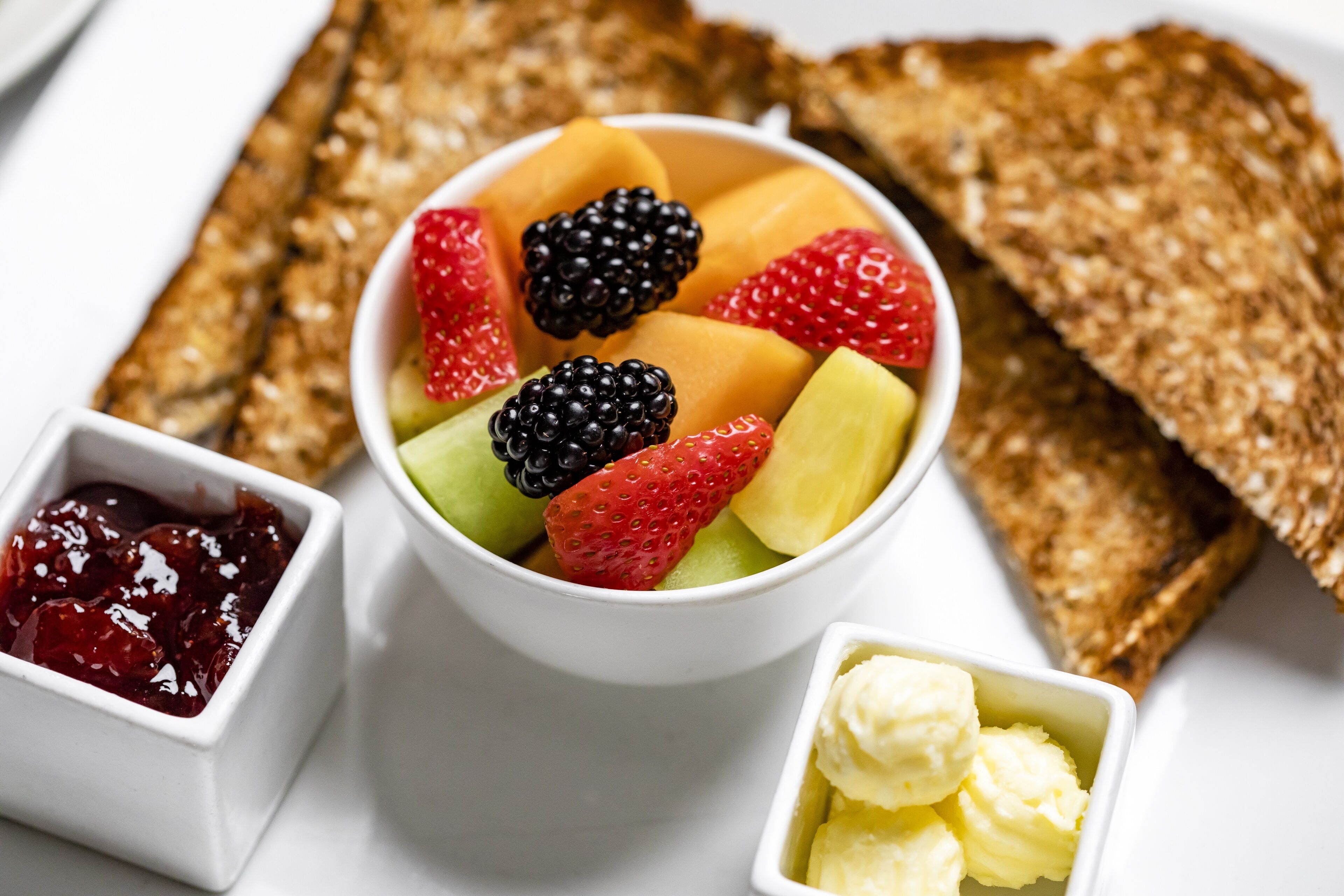 Uma saladeira com salada de frutas, morangos, amoras e melão, acompanhada de torradas e um pequeno pote de geleia.