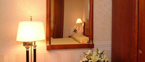 Quarto de hotel com mesa de cabeceira, luminária, espelho e arranjo floral.