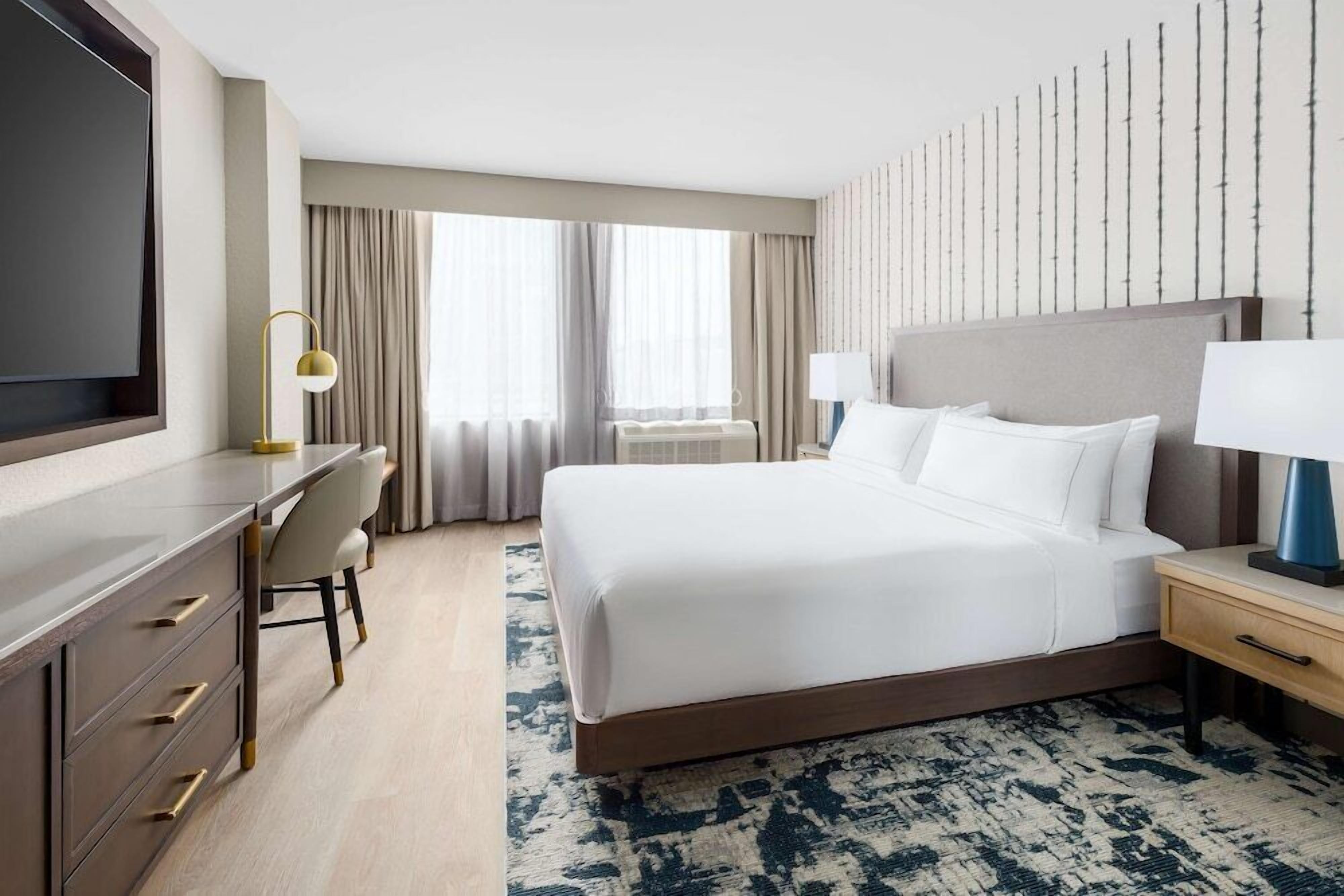 Quarto de hotel com cama, escrivaninha, televisão e janela com cortinas.
