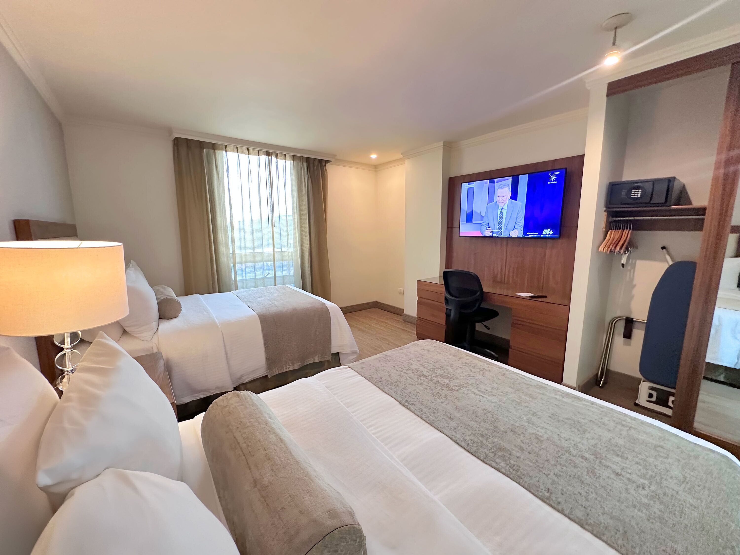 Quarto de hotel com duas camas, uma escrivaninha, uma cadeira, uma televisão e um guarda-roupa.