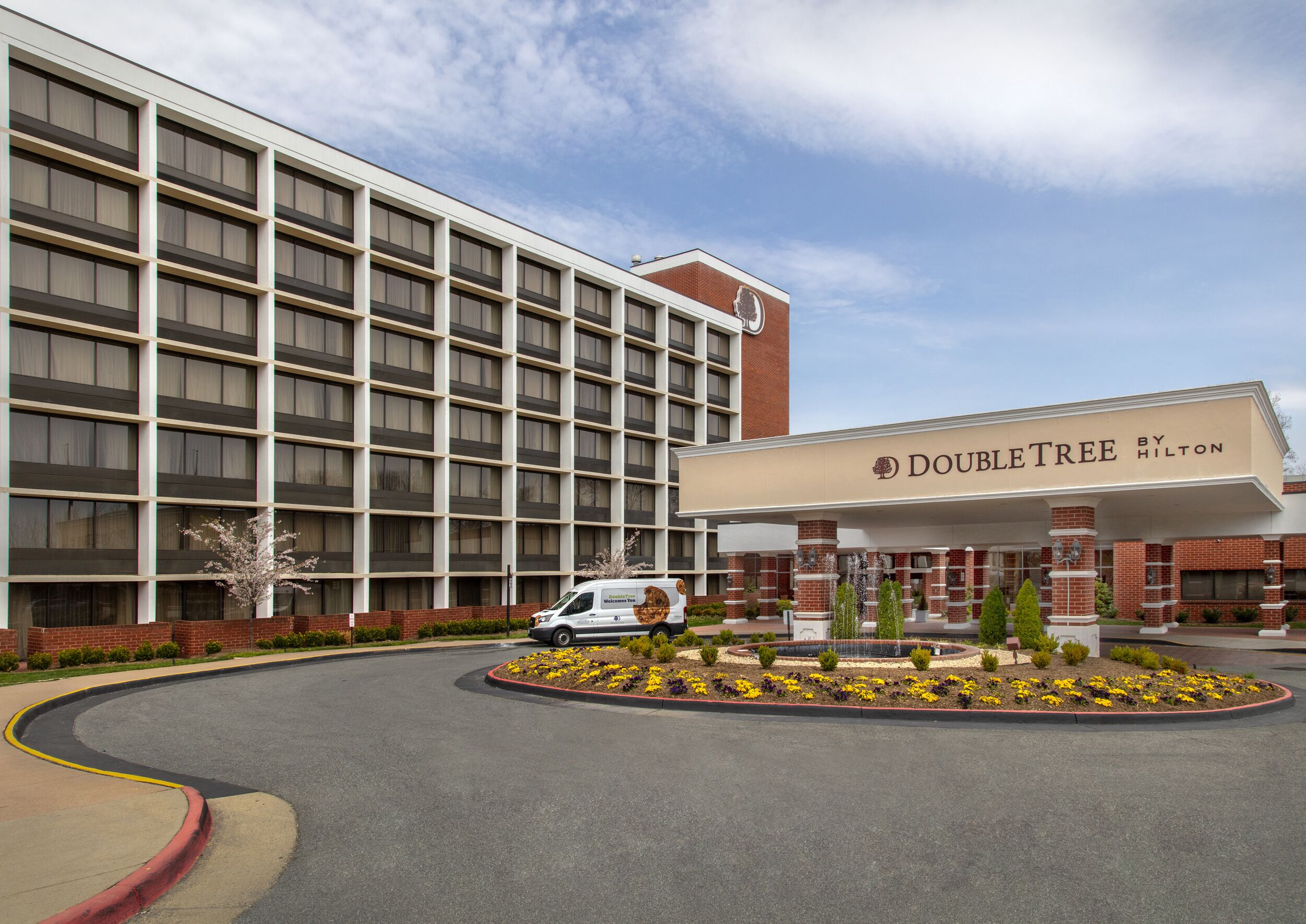 Hotel DoubleTree by Hilton com um edifício amplo, entrada convidativa e área ajardinada com flores e fonte.