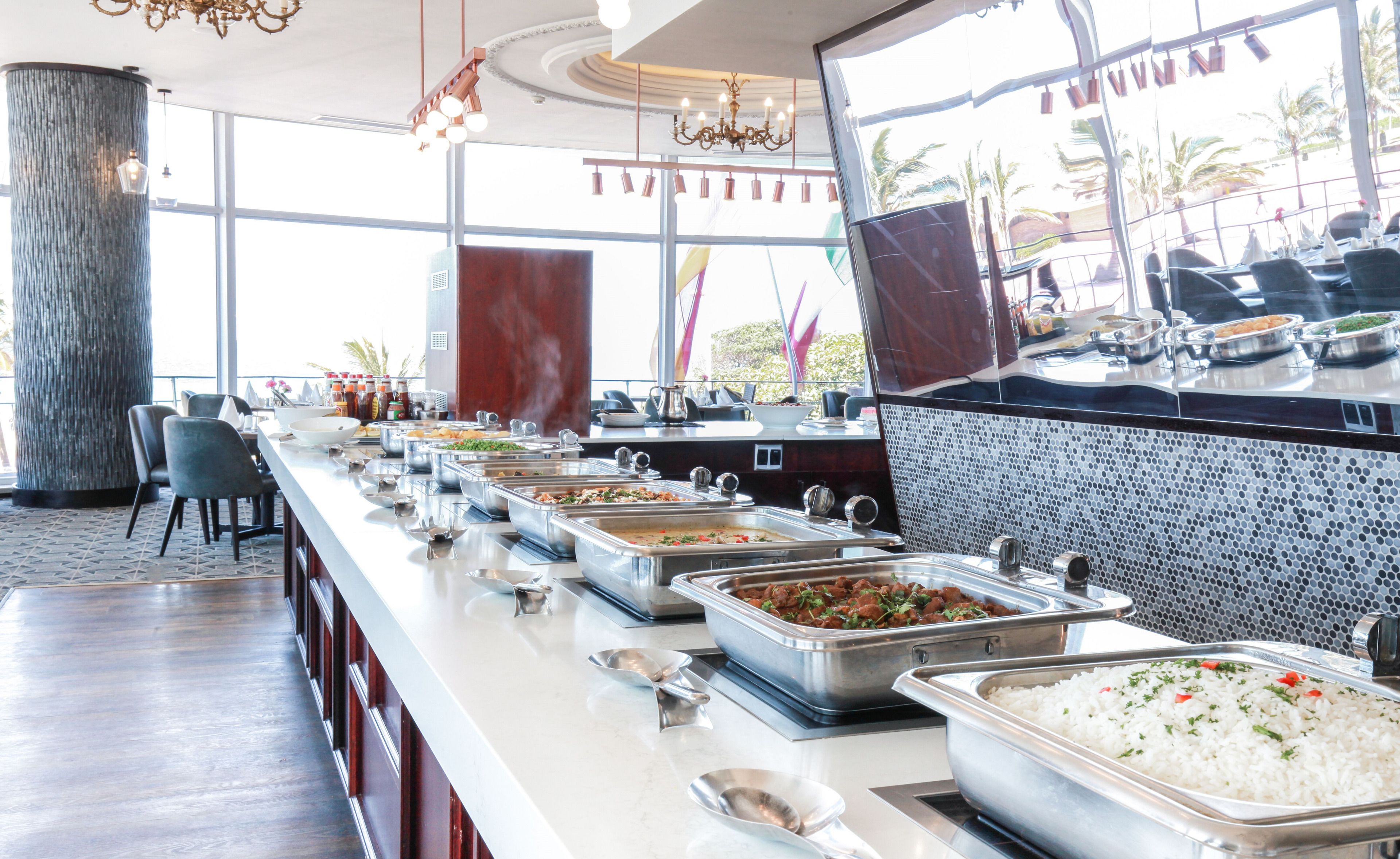 Buffet montado com diversos pontos de comida, um pilar grande e vista para palmeiras pelas janelas.