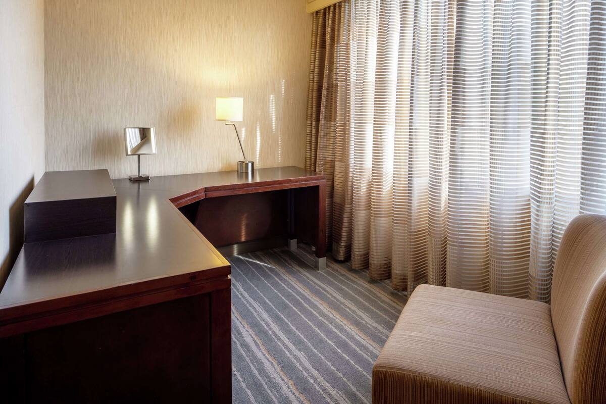 Quarto de hotel com escrivaninha, cadeira, luminária e janela com cortinas listradas.