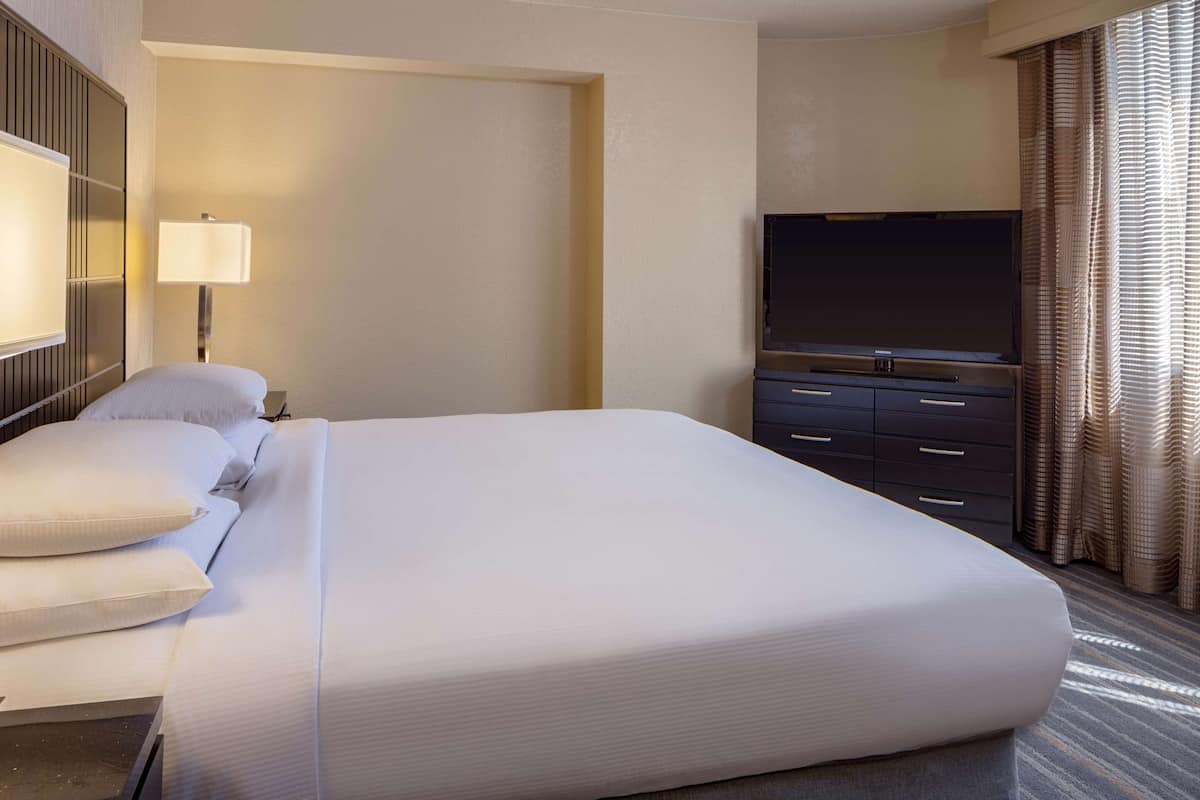 Quarto de hotel com uma cama grande, televisão, escrivaninha com abajur e armário.