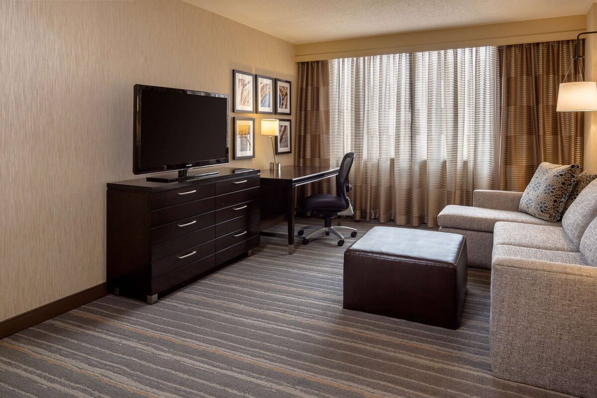 Quarto de hotel com televisão de ecrã plano, secretária, sofá confortável e uma janela grande com cortinas.