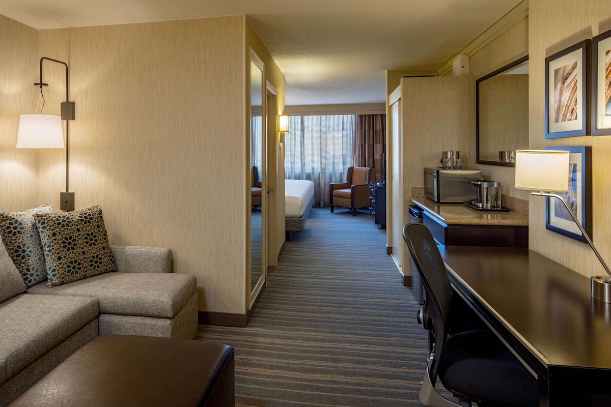 Quarto de hotel com sofá, escrivaninha, cadeira, cama e corredor que dá para outro quarto.