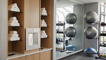 Quarto de hotel com prateleiras de madeira com toalhas enroladas, um minibar branco e um espelho refletindo equipamentos de ginástica.