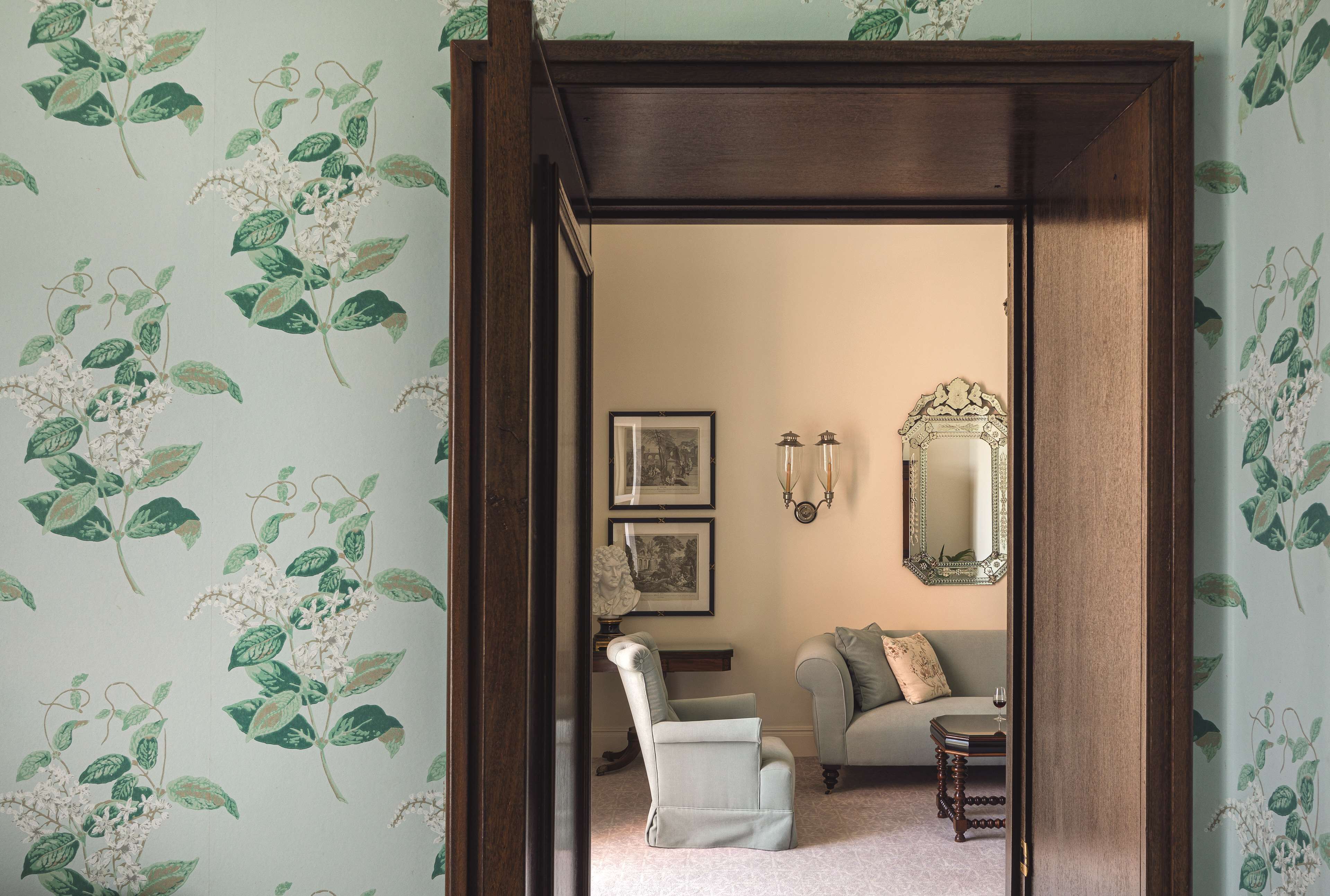 Um quarto com papel de parede floral, uma porta emoldurada em madeira, um sofá de cor clara, uma cadeira e duas imagens emolduradas na parede.