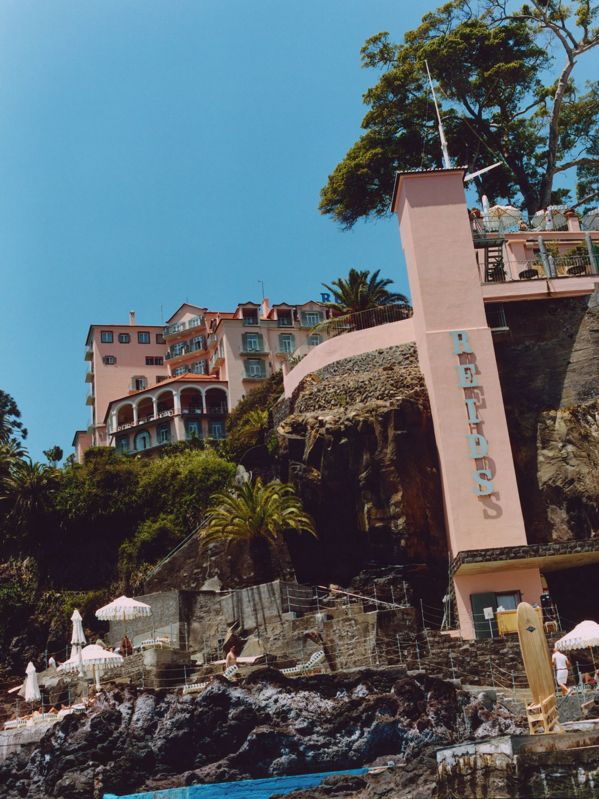 Hotel à beira-mar com uma torre rosa imponente, área de piscina com guarda-sóis e um espetacular paredão rochoso ao fundo.