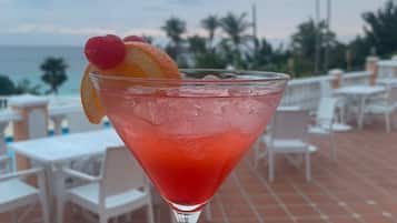 Um cocktail num copo de martini com bebida vermelha, decorado com uma fatia de laranja e um cereja, sobre uma mesa com vista para o oceano e palmeiras ao fundo.