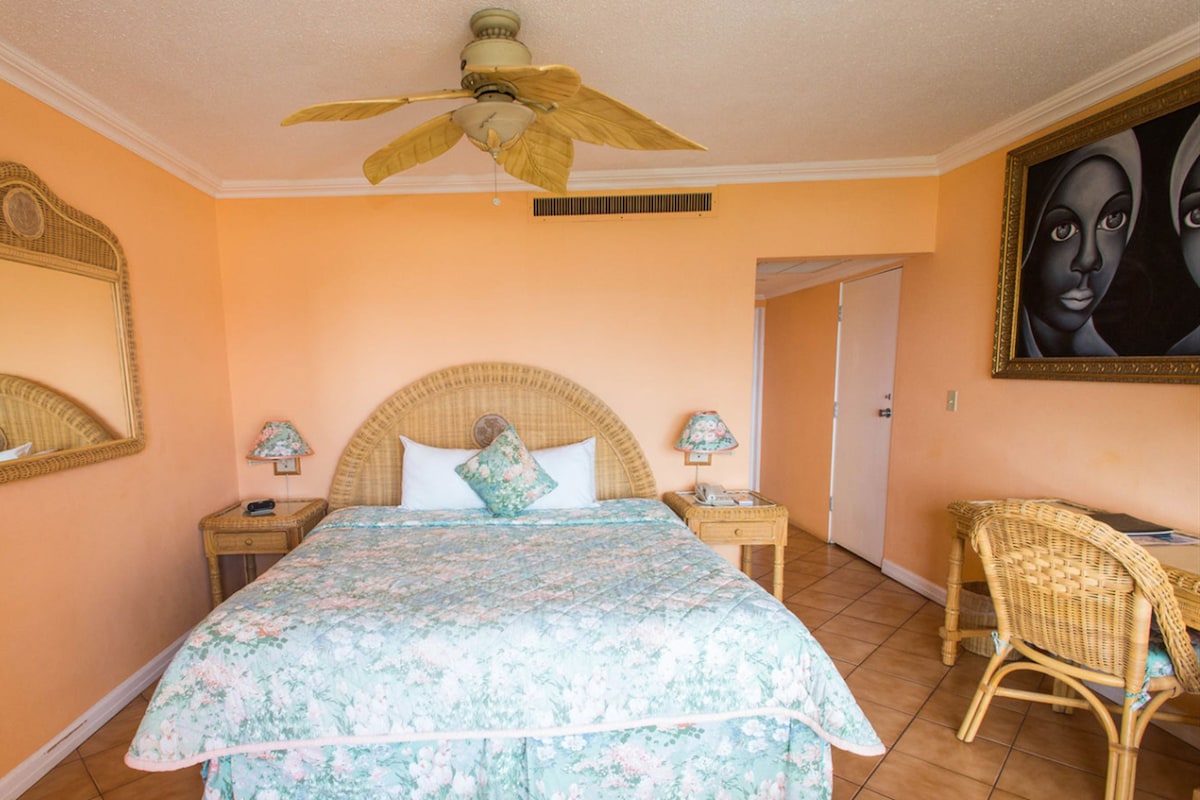 Quarto com cama, mesa de cabeceira, cadeira, ventoinha no teto, espelho e porta.