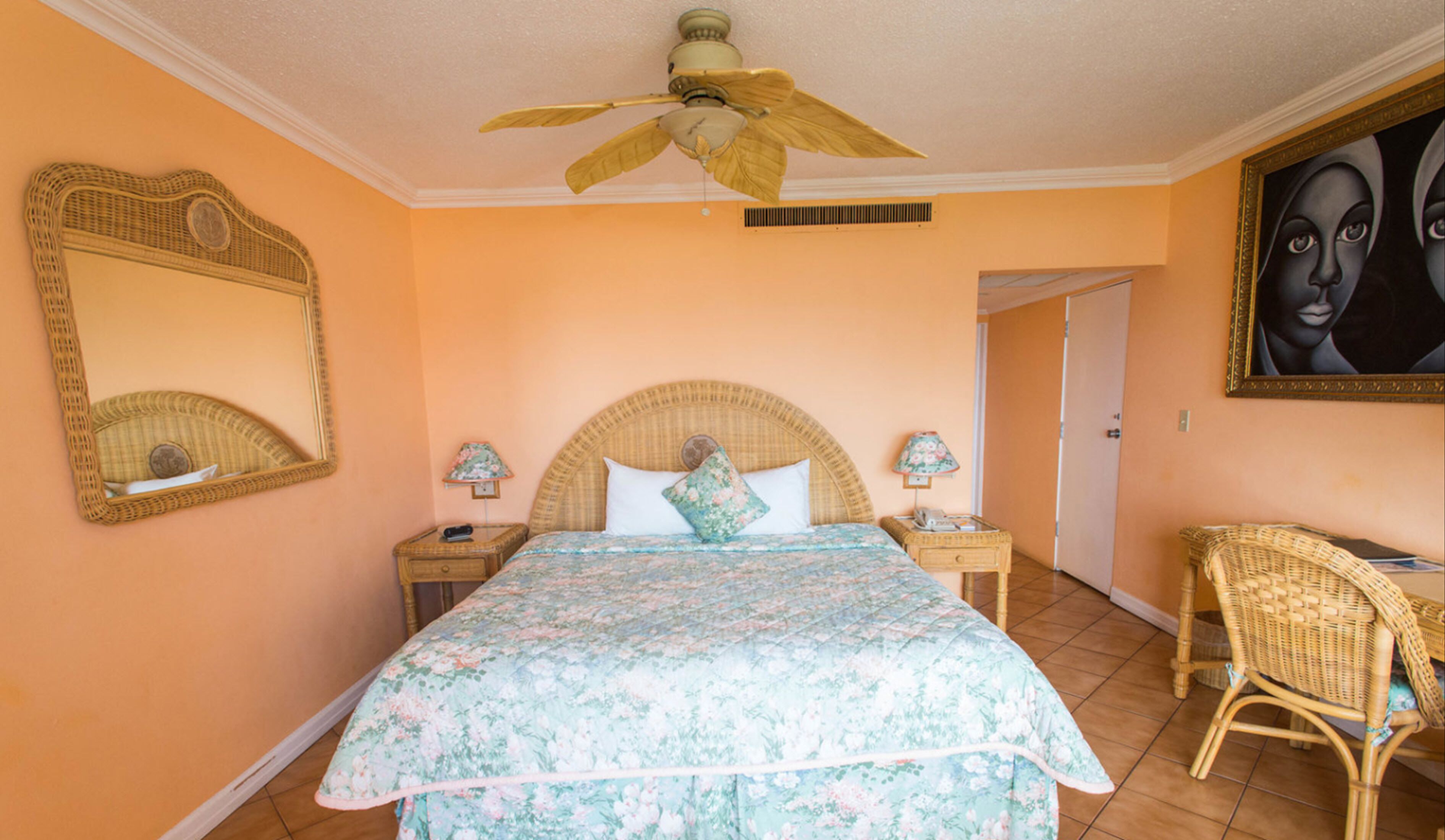 Quarto com cama, mesa de cabeceira, cadeira, ventoinha no teto, espelho e porta.