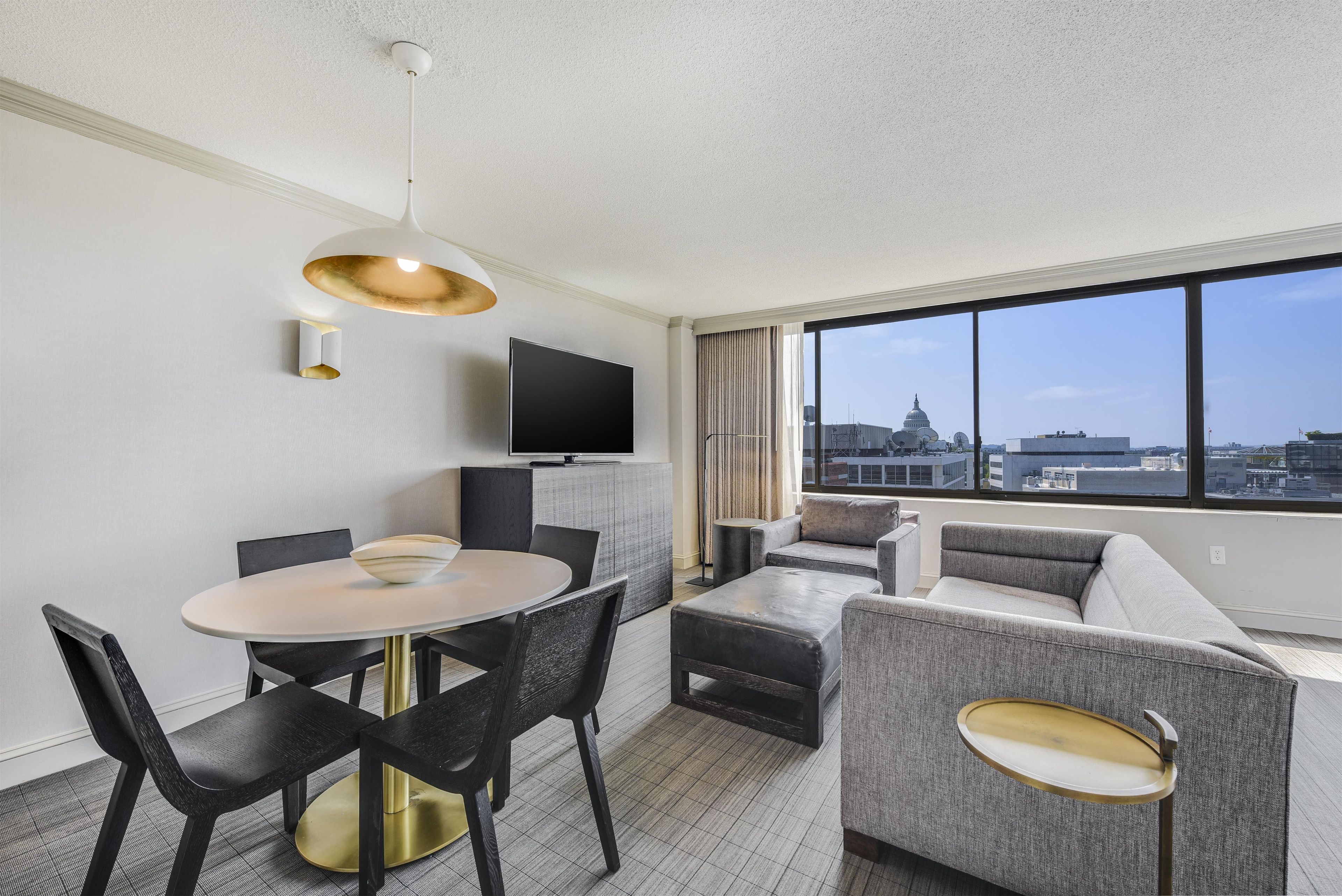 Quarto de hotel moderno com área de jantar, TV de tela plana e grande janela com vista para a cidade.