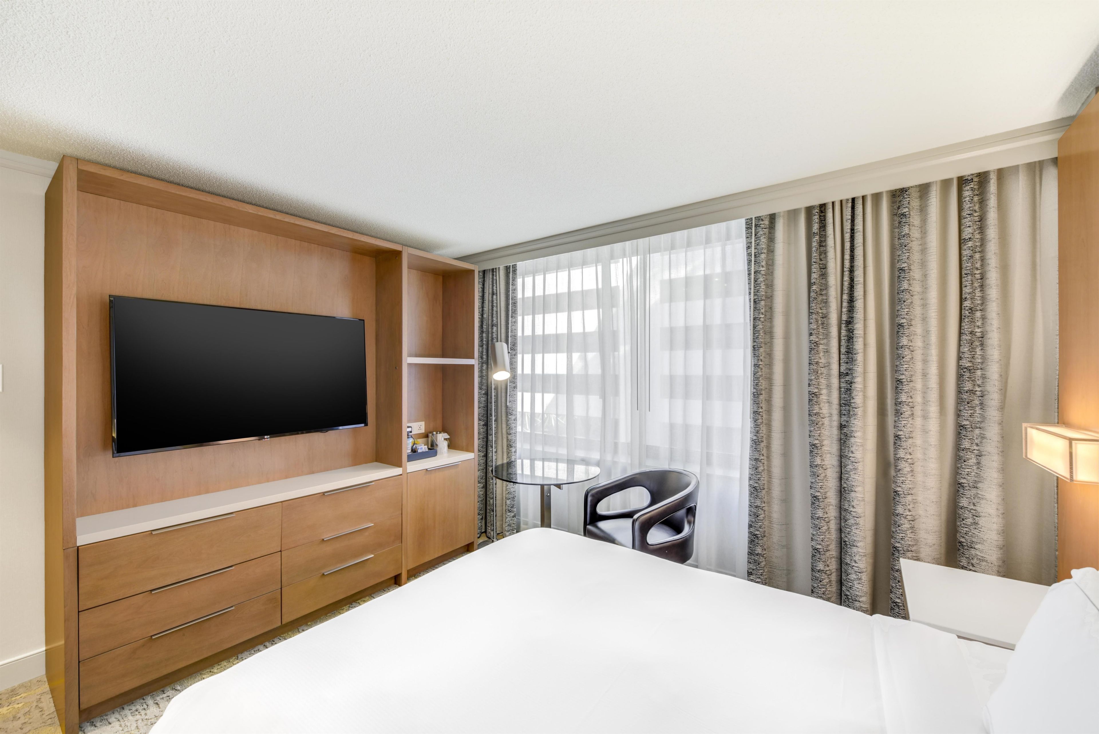 Quarto de hotel com televisão de tela plana presa em um armário de madeira, cama com roupa de cama branca, mesa redonda pequena com uma cadeira e cortinas finas com padrão discreto.