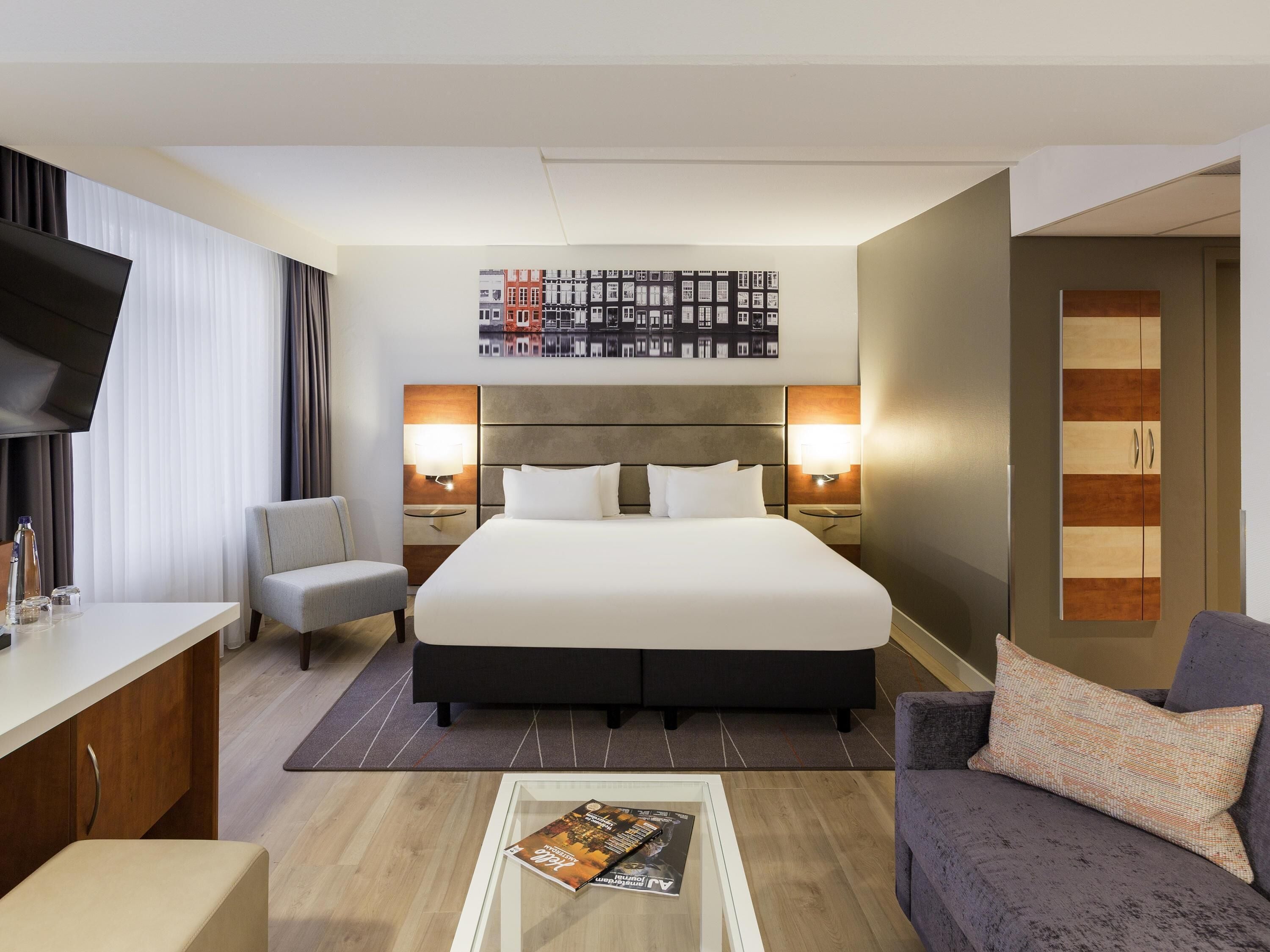 Quarto de hotel com cama grande, escrivaninha, cadeira, sofá, televisão e uma obra de arte na parede.
