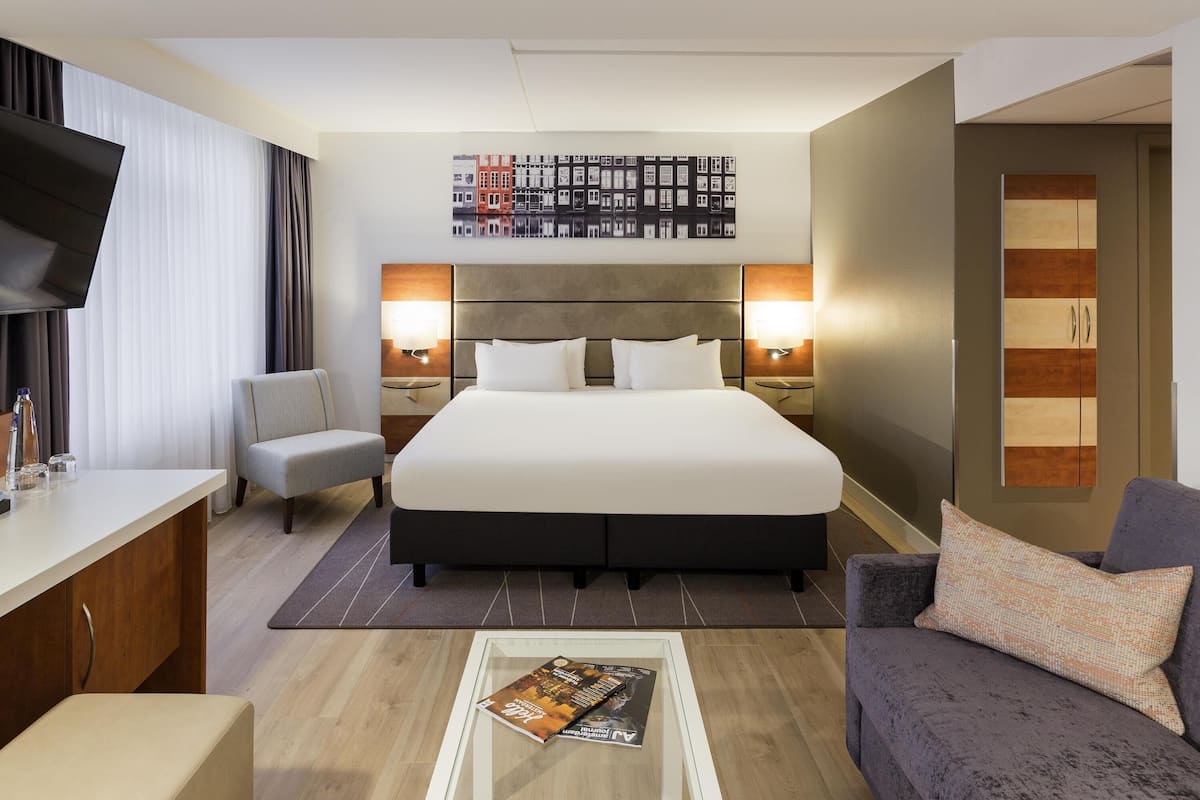 Quarto de hotel com cama grande, escrivaninha, cadeira, sofá, televisão e uma obra de arte na parede.