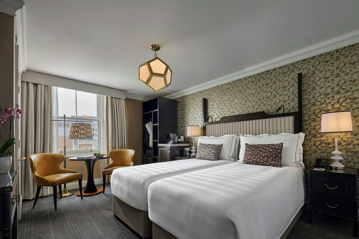 Quarto de hotel com uma cama grande, duas cadeiras, uma escrivaninha e uma janela com cortinas.