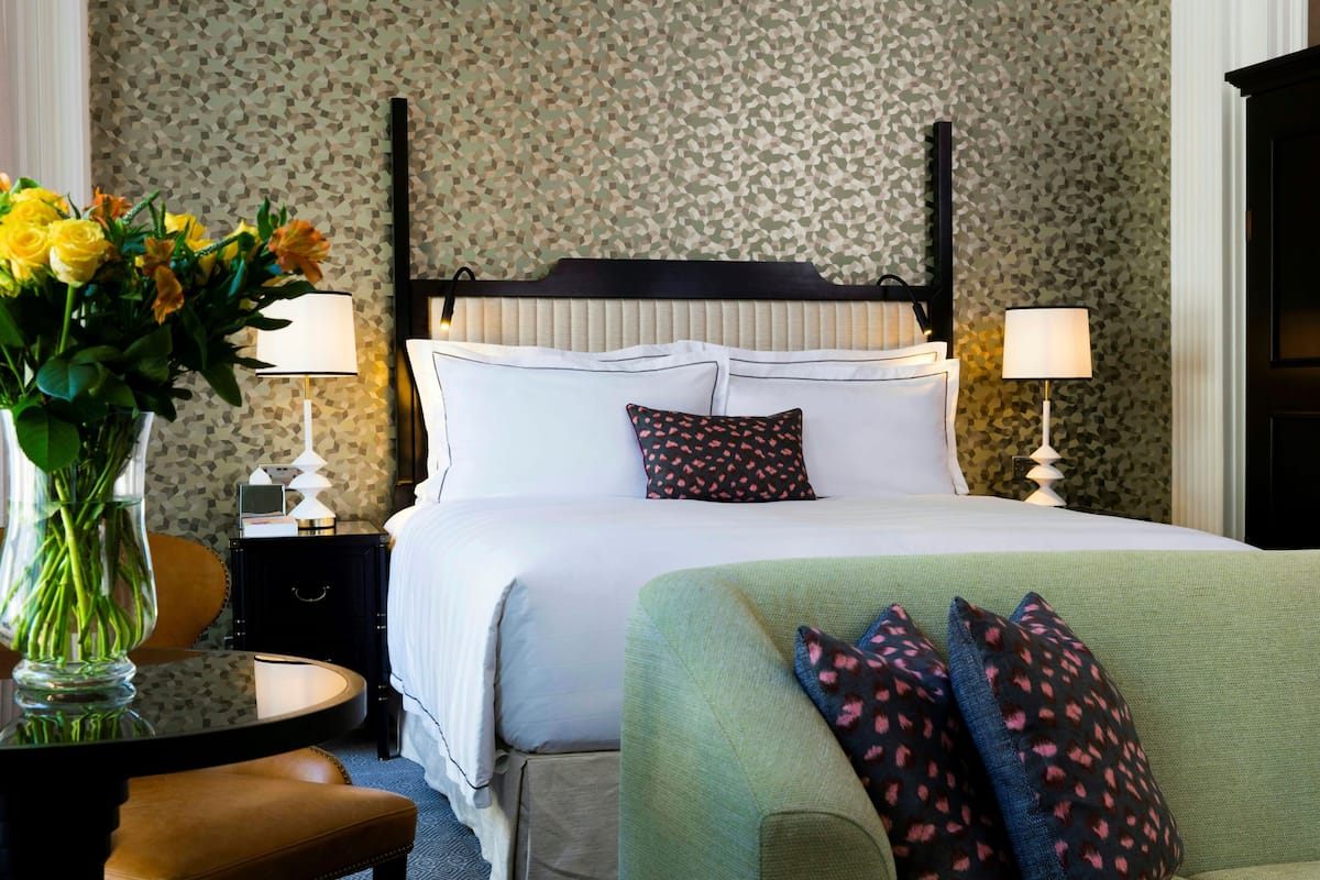 Quarto de hotel com cama de dossel, um vaso de flores, uma mesa redonda com uma cadeira e um sofá verde com almofadas estampadas.